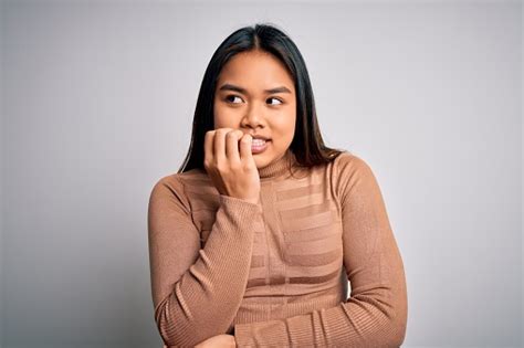 젊은 아름 다운 아시아 여자 캐주얼 터틀넥 스웨터 를 입고 흰색 배경 위에 서 스트레스와 긴장 한 입 물고 손톱에 손으로 불안 문제 갈색 머리에 대한 스톡 사진 및 기타