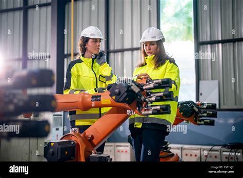 Two Female Engineers In Bright Safety Gear Discuss A Robotics Project In An Industrial Setting