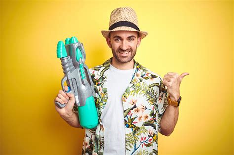 노란 고립 된 배경 가리키고 행복한 얼굴 미소와 함께 옆으로 엄지 손가락으로 보여주는 물 총을 들고 하와이 꽃 셔츠를 입고 젊은 남자 가리키기에 대한 스톡 사진 및 기타