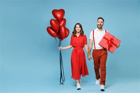 생일 휴가 파티를 축하하는 흰색 빨간 옷을 입은 전체 길이의 쾌활한 젊은 커플 친구 남자 여자는 파란 배경에 고립 된 선물로 헬륨 풍선 선물 상자를 들고 있습니다 2명에