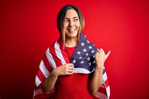 젊은 아름다운 금발의 애국적인 여자 와 푸른 눈을 입고 미국 국기 를 가리키고 행복한 얼굴 미소와 함께 측면에 엄지 손가락으로 보여주는 가리키기에 대한 스톡 사진 및 기타