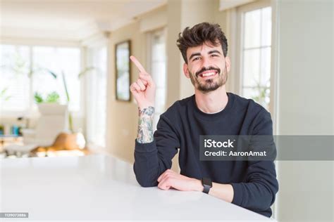 젊은 남자가 얼굴에 큰 미소로 흰색 테이블에 앉아 캐주얼 셔츠를 입고 카메라를 보고 측면에 손과 손가락을 가리키는 가리키기에 대한 스톡 사진 및 기타 이미지 Istock
