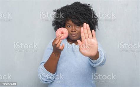 심각 하 고 자신감 표현 방어 제스처 정지 표시를 하 고 오픈 손으로 젊은 아프리카계 미국인 여자 회색 그런 지 벽 먹는 동안 핑크 도넛 경계 표지에 대한 스톡 사진 및