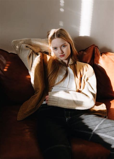 햇살이 잘 드는 방에서 가죽 소파에 앉아 휴식을 취하는 젊은 여성 · 무료 스톡 사진