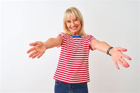 캐주얼 한 줄무늬 티셔츠를 입은 중년 여성이 고립 된 흰색 배경 위에 서서 포옹을 위해 팔을 벌려 미소짓고있는 카메라를 바라보고 있습니다 행복을 포용하는 쾌활한 표현 팔