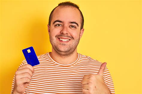 확인 기호를하고 큰 미소행복 고립 된 노란색 배경 위에 서 파란색 신용 카드를 들고 젊은 남자 손가락으로 엄지 손가락 우수한 기호 고객에 대한 스톡 사진 및 기타 이미지