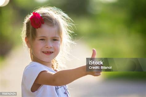 흐릿한 밝은 녹색 여름 배경에 카메라에 엄지 손가락 제스처를 보여주는 머리에 빨간 장미와 흰색 티셔츠에 귀여운 작은 금발 심각한 소녀의 흐릿한 초상화 어린 시절 개념의