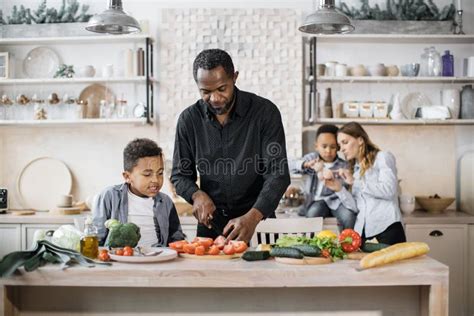 Handsome African Father And Son Preparing Tasty Salad At Kitchen While Choping Vegetables Stock