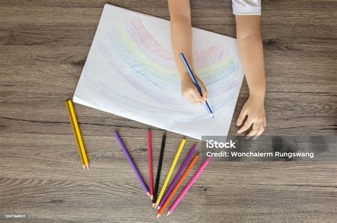 위에서 찍은 사진은 미취학 아동이 나무 테이블에 놓인 흰색 종이에 컬러 연필로 그려진 다채로운 무지개 드로잉을 위해 창의력과 즐거움을 개발하는 법을 배우는 두 팔을 볼 수