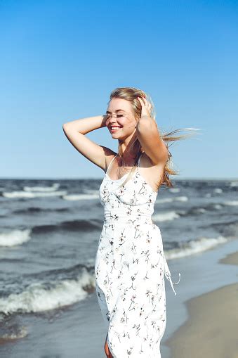 바다 해변에서 자유로운 행복의 행복을 누리고 있는 행복한 금발 여자가 똑바로 서 있다 흰색 여름 드레스를 입은 여성 모델의 초상화는 야외에서 휴가 휴가 동안 자연을 즐기고