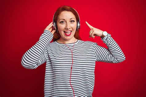 젊은 빨간 머리 여자 듣고 음악을 사용 하 여 헤드폰 을 통해 빨간 고립 된 배경 매우 행복 가리키는 손과 손가락 가리키기에 대한 스톡 사진 및 기타 이미지 Istock