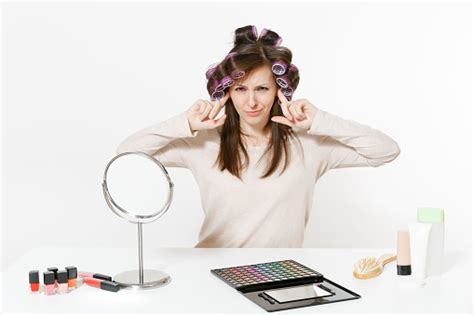 Curlers 재미 여자는 흰색 바탕에 격리 설정된 얼굴 장식 화장품으로 메이크업을 적용 하는 테이블에 앉아 손으로 귀를