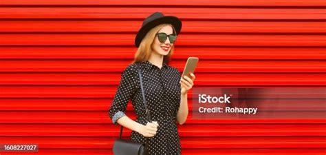 검은색 둥근 모자를 쓰고 스마트폰을 들고 빨간 배경에 핸드백을 들고 있는 세련된 웃는 여자의 초상화 20 29세에 대한 스톡 사진 및 기타 이미지 Istock