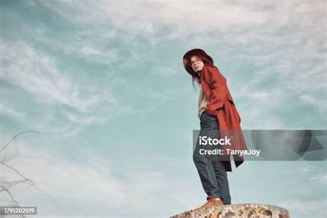 빨간 코트와 모자를 쓴 자신감 넘치는 십대 소녀가 주머니에 손을 넣고 하늘을 배경으로 서 있습니다 로우 앵글 뷰 14 15 살에 대한 스톡 사진 및 기타 이미지 Istock