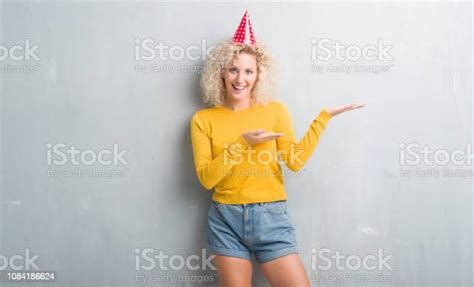 그런 지 회색 벽 입고 생일에 젊은 금발의 여 자가 깜짝 놀라게 하 고 카메라에 웃는 동안 손으로 제시와 함께 손가락 포인팅 모자 가리키기에 대한 스톡 사진 및 기타 이미지