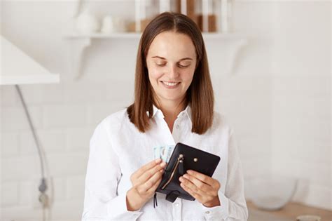 흰색 캐주얼 스타일 셔츠를 입은 부유한 여성의 실내 샷 손에 검은 지갑과 함께 가벼운 부엌에서 포즈 쇼핑을 가기 전에 그녀의 저축을 계산 지갑 개인 장식품에 대한 스톡 사진