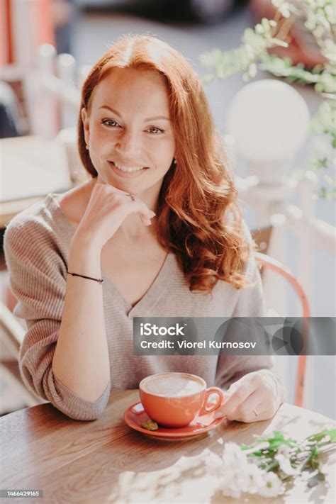 사람과 자유 시간 개념입니다 부드러운 미소와 함께 긍정적인 빨간 머리 여자 턱 아래 손을 유지 캐주얼 점퍼를 입고 음료 향기로운 커피 야외 카페테리아에서 친구와 이야기를