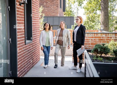 Redhead Realtor In Glasses Holding Folder While Looking At New House Near Cheerful Interracial