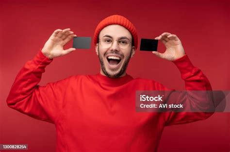 안경과 빨간 옷을 입은 수염을 가진 매력적인 남자의 사진 행복한 남자는 빨간색 배경에 고립 된 2 신용 카드를 보유하고 있습니다 25 29세에 대한 스톡 사진 및 기타