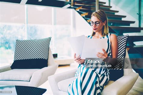 세련된 베개와 편안한 소파에 앉아 모의 영역안경 확인 종이 문서에 똑똑한 젊은 여성 펜션 여성 편집 과정 작업 휴식 에 소파 에 현대 홈 인테리어 계약서에 대한 스톡 사진