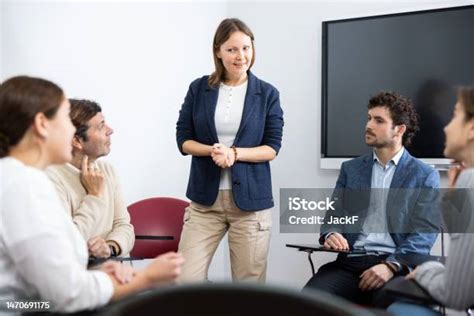 중년 여성 비즈니스 코치는 강당에서 원형으로 앉아 있는 사람들의 그룹에 대해 인터랙티브 보드 근처에 서서 손으로 몸짓을 하고