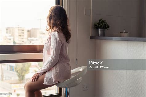 분홍색 셔츠에 아름 다운 혼자 갈색 머리 여자 높은 의자에 앉아 그녀의 아파트의 거 대 한 창 밖으로 보이는 가정에서 외로움과 고립의 개념 가정 주방에 대한 스톡 사진 및