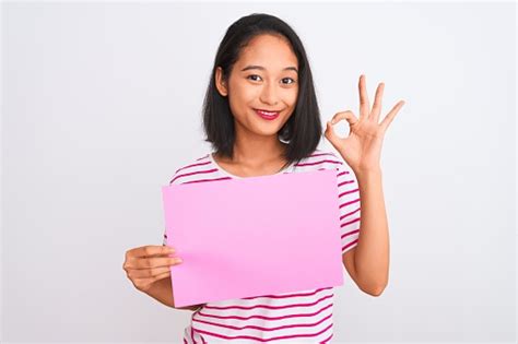 손가락으로 확인 기호를하고 고립 된 흰색 배경 위에 서있는 배너를 들고 젊은 아름다운 중국 여자 우수한 기호 개념에 대한 스톡