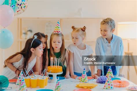 작은 아이들은 생일 파티를 축하하고 케이크에 촛불을 불어 축제 테이블에 모여 좋은 분위기를 가지고 함께 시간을 보내고 소원을 만들고 파티 모자를 쓰고 팽창 된 풍선으로 실내