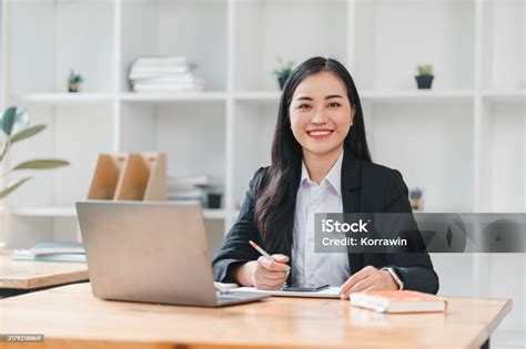 자신감 넘치는 여성 사업가가 책상에서 일하면서 미소를 지으며 현대적인 사무실 환경을 보여줍니다 그녀는 노트북과 노트에 몰두하여 전문성과 생산성을 반영합니다 25 29세에