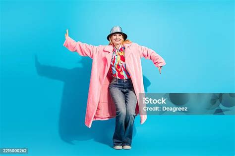 화려한 의상을 입고 금발 머리를 한 세련된 성숙한 여성이 현대적인 패션을 보여주는 파란색 배경에 활기차게 포즈를 취하고 있습니다 50 54세에 대한 스톡 사진 및 기타