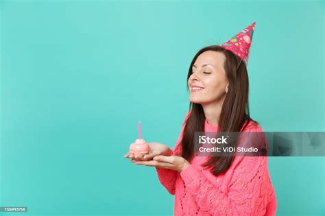 젊은 여자 니트 핑크 스웨터에 웃 고 닫힌된 눈으로 생일 모자 손에 촛불 블루 벽 배경 스튜디오 초상화에 고립 된 케이크를 개최 사람들의 라이프 스타일 개념입니다 복사