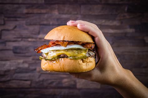 치즈 계란 베이컨 피클을 나무 테이블과 소박한 배경에 얹은 수제 소고기 버거를 손에 들고 있습니다 고기에 대한 스톡 사진 및 기타 이미지 Istock