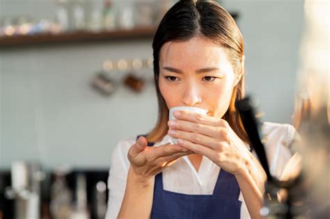 자신감 넘치는 아시아 일본 여성 또는 바리스타가 앞치마를 입고 커피숍 카운터 바에 서서 햇빛 아래 미소로 무장하고 흰색 컵에서 블랙 커피 냄새를 맡고 있습니다 뜨거운 음료의