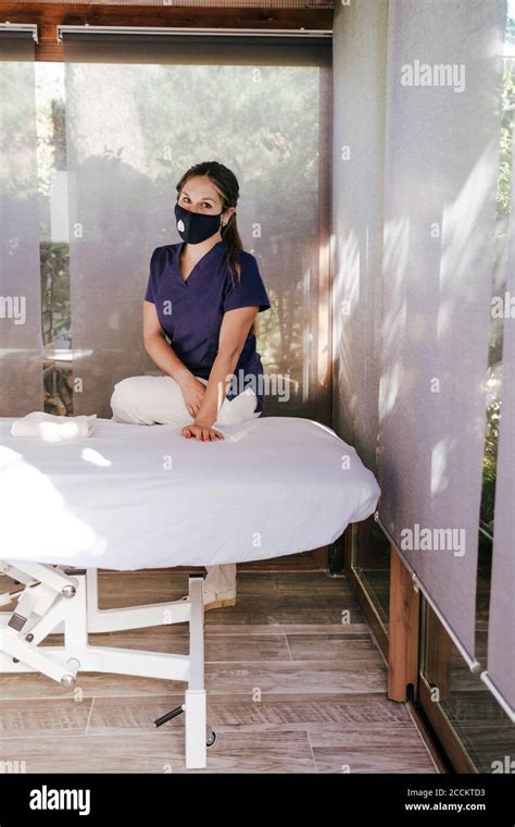 Female Therapist Wearing Mask Standing By Massage Table In Spa Stock