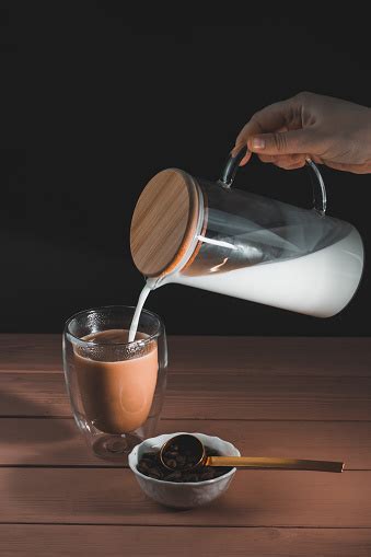 소녀는 이중 벽유리 컵에 우유를 붓습니다 우유 디켄터 나무 테이블과 검은 색 테이블 흰색 접시에 커피 콩 갈색에 대한 스톡 사진