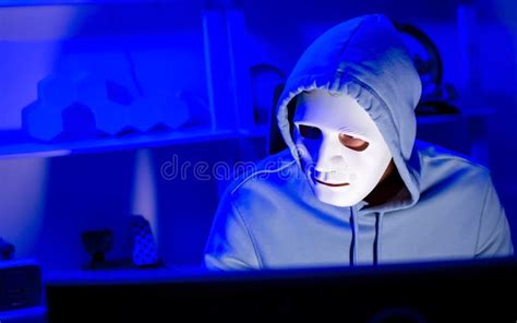 Male Hacker Wearing Mask And Hoodie Sitting In Dark Room Looking At Camera Making Cyber