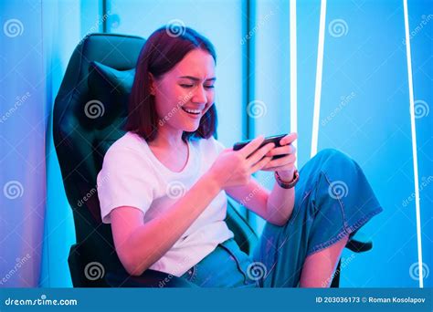 Cute Gamer Girl Sit On A Gaming Chair Smiling And Playing Mobile Online Game On A Smartphone