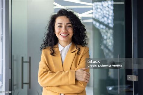 사무실 안에서 성공한 여성 사업가의 초상화 노란 양복을 입고 웃으며 카메라를 바라보는 라틴 아메리카 상사 팔짱을 끼고 서 있는 곱슬머리를 한 성숙한 여성 노동자 노랑에 대한