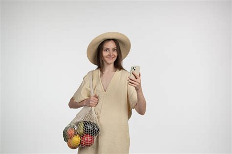 베이지색 의상을 입은 젊은 여성이 실내에 서서 형형색색의 과일이 가득 담긴 망사 가방을 들고 휴대전화를 보며 미소를 짓고 있다 은은한 조명이 밝은 분위기를 돋보이게 합니다