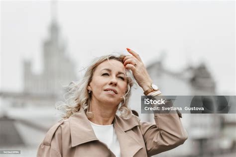 곱슬 머리를 가진 중년의 금발 여성이 도시의 강 제방에 서 있습니다 길거리 스타일에 대한 스톡 사진 및 기타 이미지 길거리 스타일 여자 바람 Istock