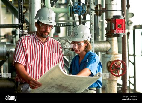 Male And Female Process Engineers Wearing Appropriate Safety Equipment Consult A Blueprint On A
