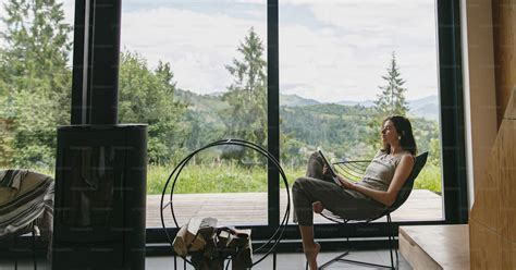 산 언덕을 배경으로 장작이 있는 벽난로 의자에서 책을 읽는 아름다운 세련된 여성 창문에서 놀라운 전망을 감상할 수 있는 현대적인 샬레에서 편안한 캐주얼 옷을 입은 젊은