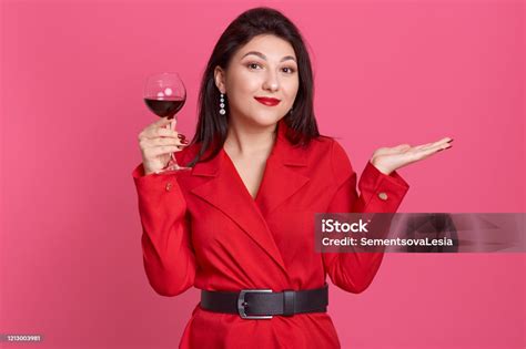 와인 한 잔을 들고 손을 벌리고 손을 벌리고 갈색 머리 여성이 카메라를 직접 보고 핑크 스튜디오 배경 위에 고립 된 포즈를 취하고 생일을 축하하는 여성의 스튜디오 샷