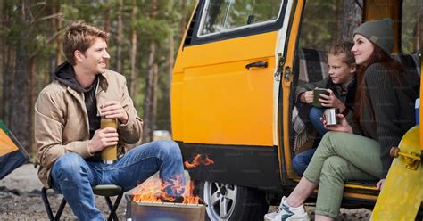 웃는 아버지는 차와 함께 보온병을 열고 밝은 태양을 향해 곁눈질합니다 자연에 앉아 아늑한 숲의 캠프 파이어에서 컵으로 뜨거운 차를 마시는 행복한 가족 사진