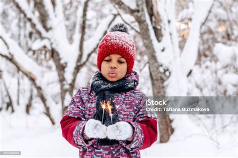 빨간 모자와 점프 슈트를 입은 작은 슬픈 아프리카 계 미국인 소녀는 겨울 숲에서 불타는 스파클러를 손에 쥐고 있습니다 겨울 재미 메리 크리스마스와 새해 복 많이 받으세요