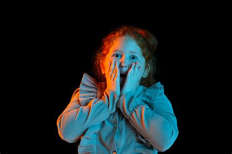 충격 네온 빛으로 어두운 배경에 고립 된 축제 드레스를 입은 귀여운 빨간 머리 어린 소녀의 절반 길이의 초상화 어린이 감정 얼굴