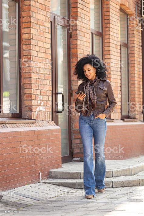 그녀의 스마트 폰에 문자 메시지 젊은 아프리카 계 미국인 여성의 초상화 가죽 재킷과 세련된 청바지에 곱슬 머리와 갈색 머리 는 흐린 벽돌 건물의 배경으로 거리에서 포즈 갈색