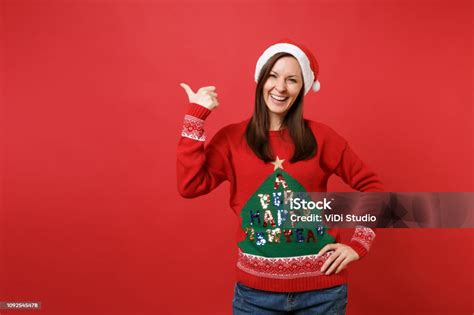 니트 스웨터와 크리스마스 모자 옆으로 밝은 빨간 벽 배경에 고립 된 엄지를 가리키는 산타 소녀 웃 고 있다 행복 한 새 해 2019 축 하 휴일 파티 개념입니다 복사 공간을