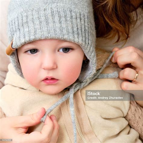 어머니는 집 복도에 앉아있는 유아 아기의 머리에 따뜻한 모자를 씌 웁니다 여자 엄마는 추운 날씨에 겨울 산책을 위해 아이에게 따뜻한 옷을 입는다 1세 3개월 된 아이 매기에