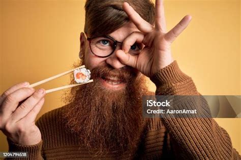 빨간 머리 아일랜드 사람 수염 먹는 연어 마키 초밥 노란 배경 위에 젓가락을 사용 하 여 행복 한 얼굴 웃고 손가락으로 보고 눈으로 확인 기호를 하 고 유머에 대한 스톡